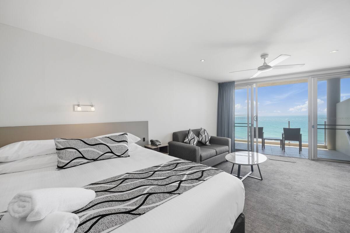 Quarto de hotel com cama, sofá, mesa pequena e varanda com vista para o mar.