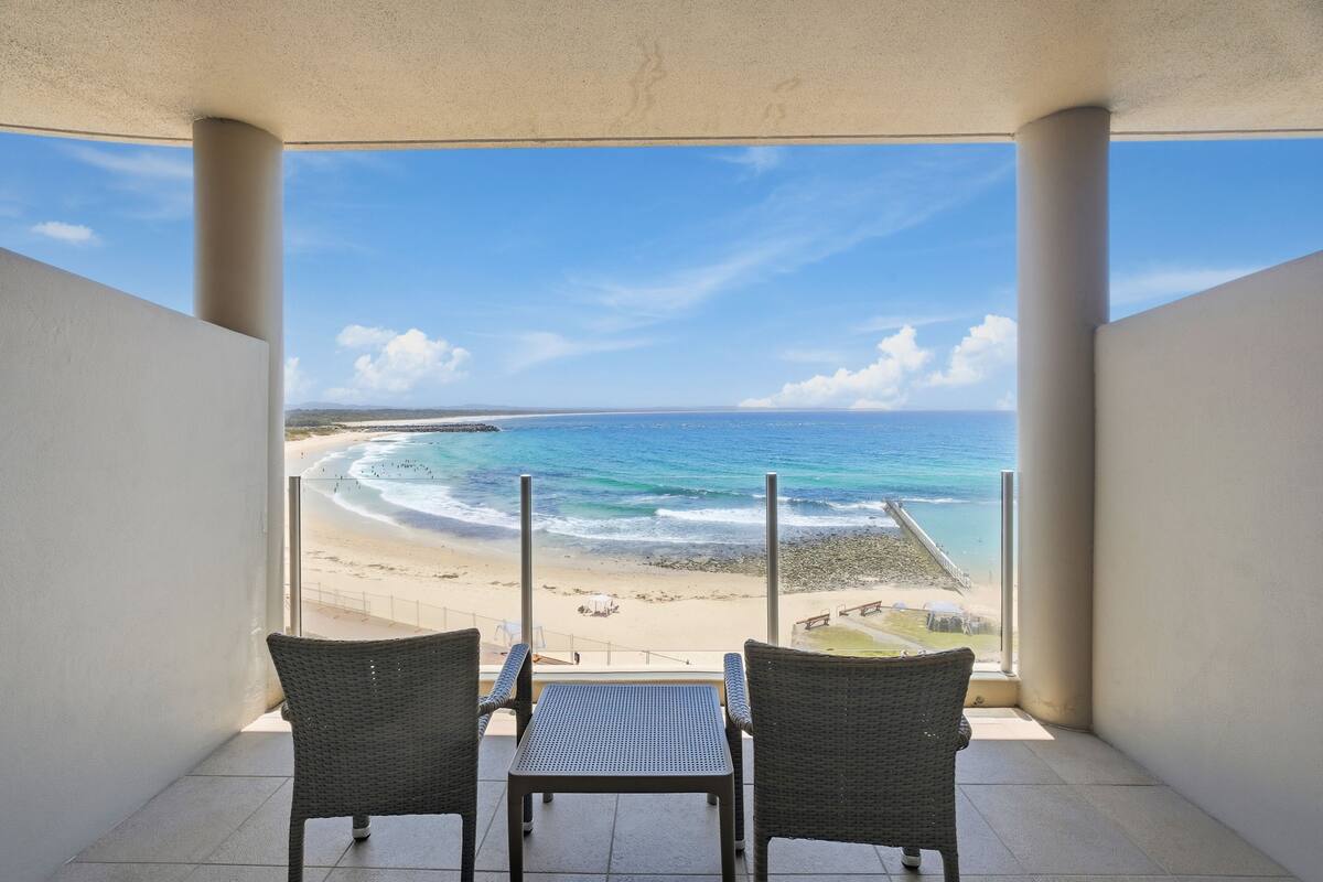 Varanda com duas cadeiras de vime e uma mesa, com vista para uma praia com ondas e céu claro.