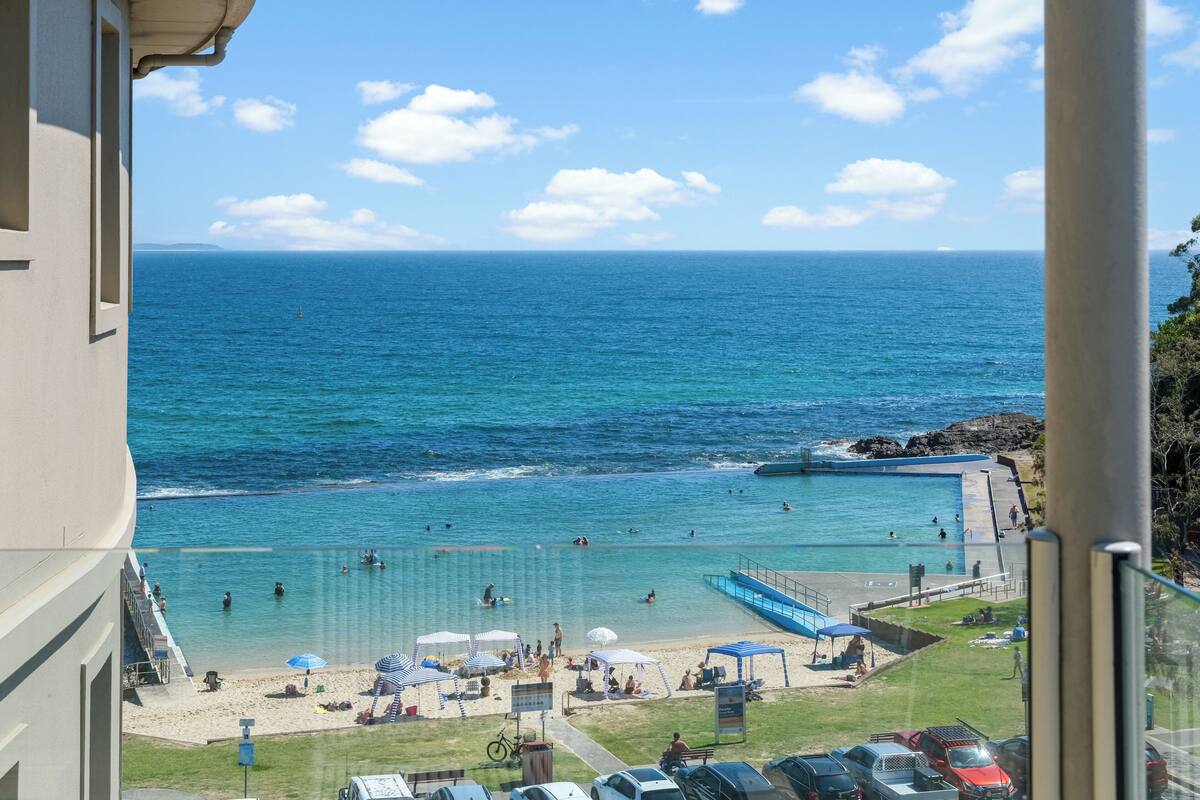 Vista de uma praia com piscina, guarda-sóis e carros estacionados na lateral.