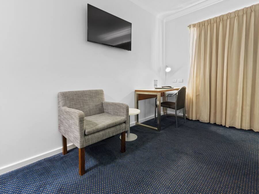 Quarto de hotel com carpete azul, uma poltrona cinza, escrivaninha de madeira, televisão de tela plana na parede e janela com cortinas bege.