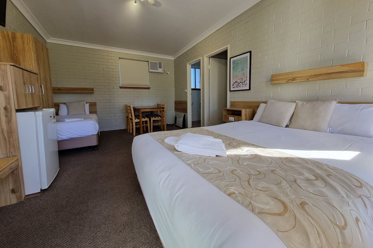 Quarto de hotel com uma cama grande, uma cama menor, uma escrivaninha e um frigorífico.