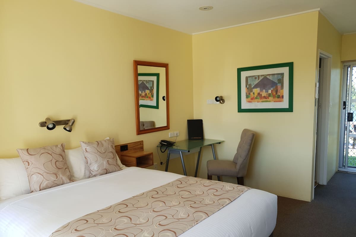 Quarto de hotel com uma cama grande, uma escrivaninha, uma cadeira, um espelho e duas imagens emolduradas na parede.