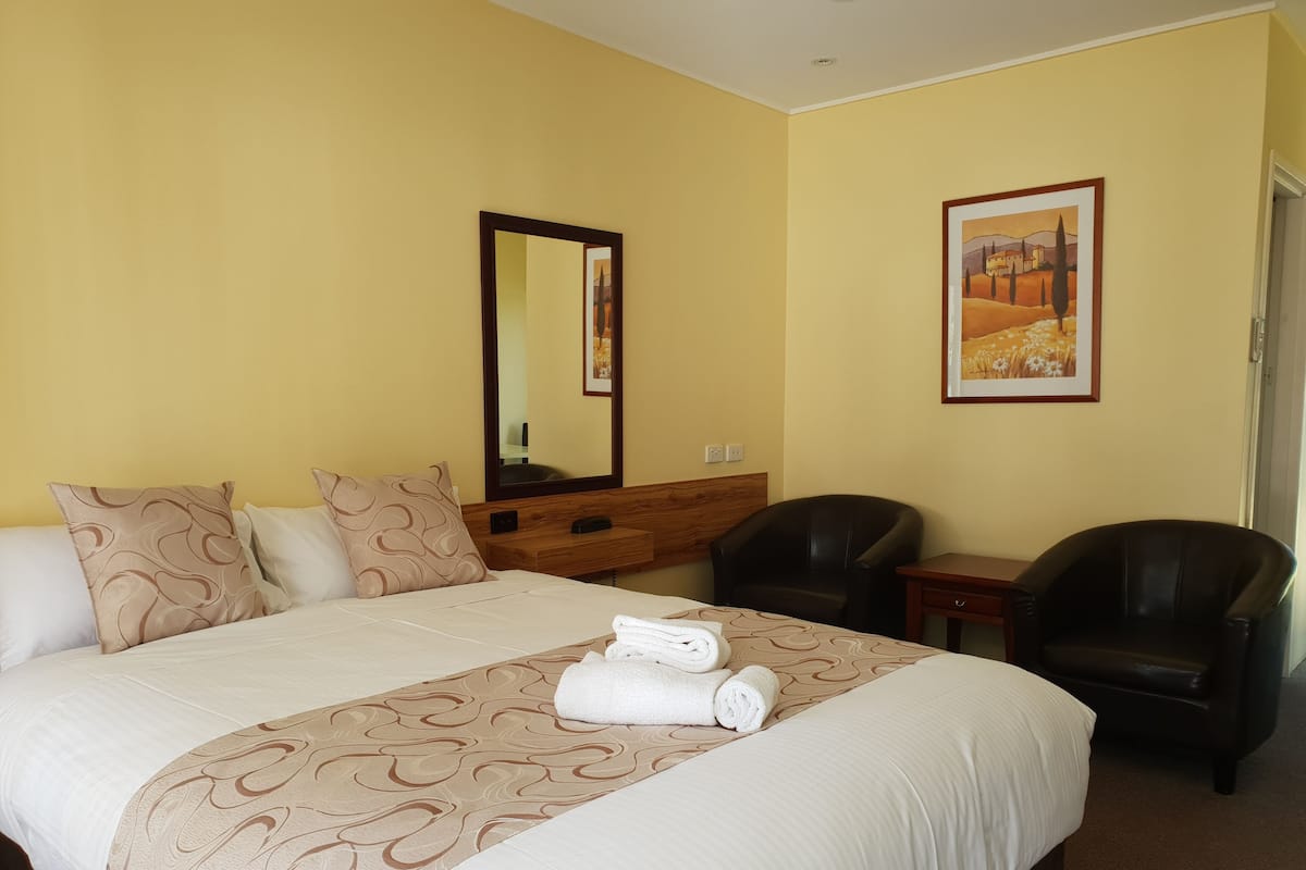 Quarto de hotel com uma cama grande, duas cadeiras, uma mesa de cabeceira de madeira, um espelho e um quadro na parede.