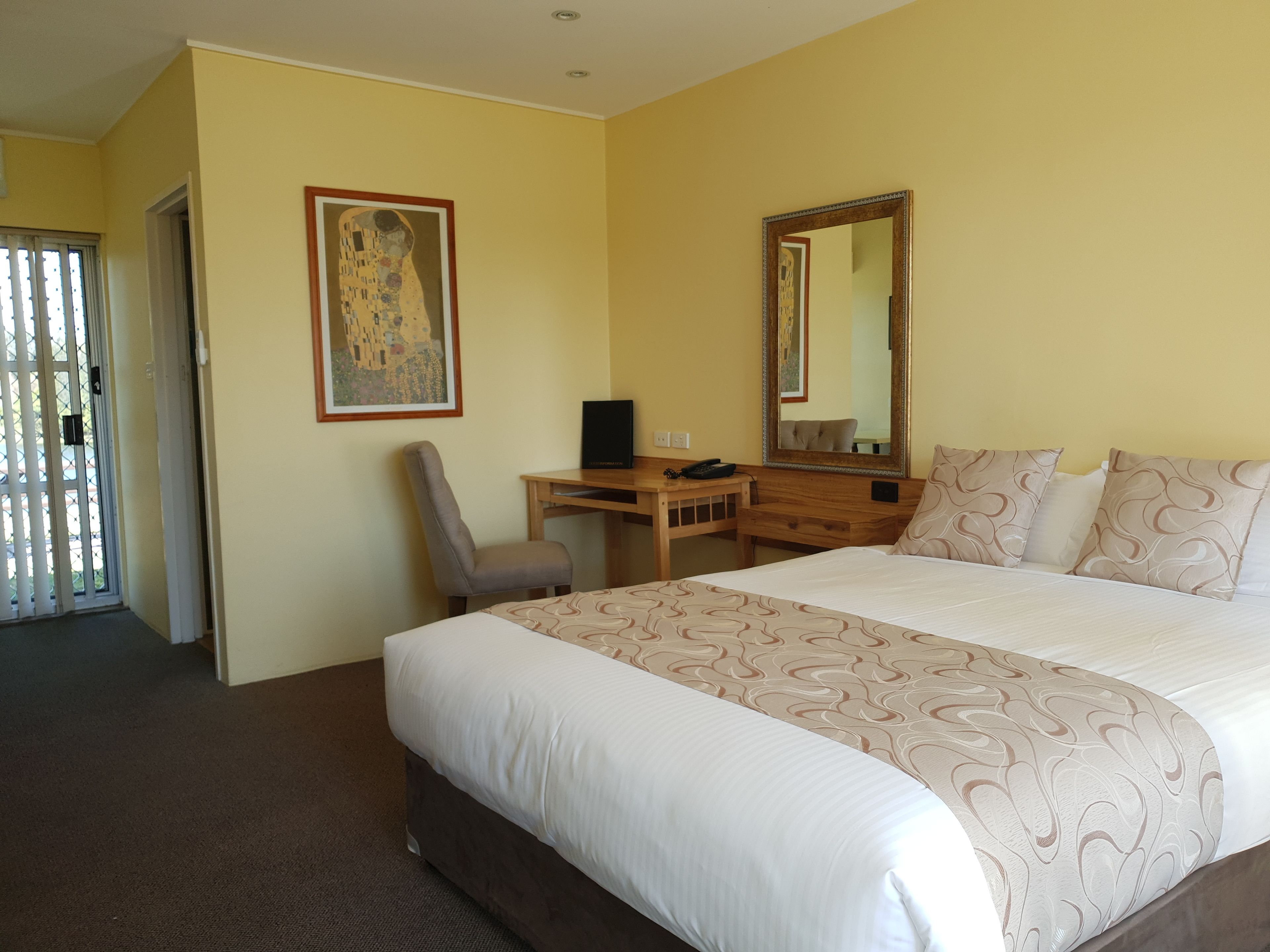 Quarto de hotel com uma cama grande, uma escrivaninha com cadeira, luminária de mesa, telefone, quadro emoldurado e um espelho.