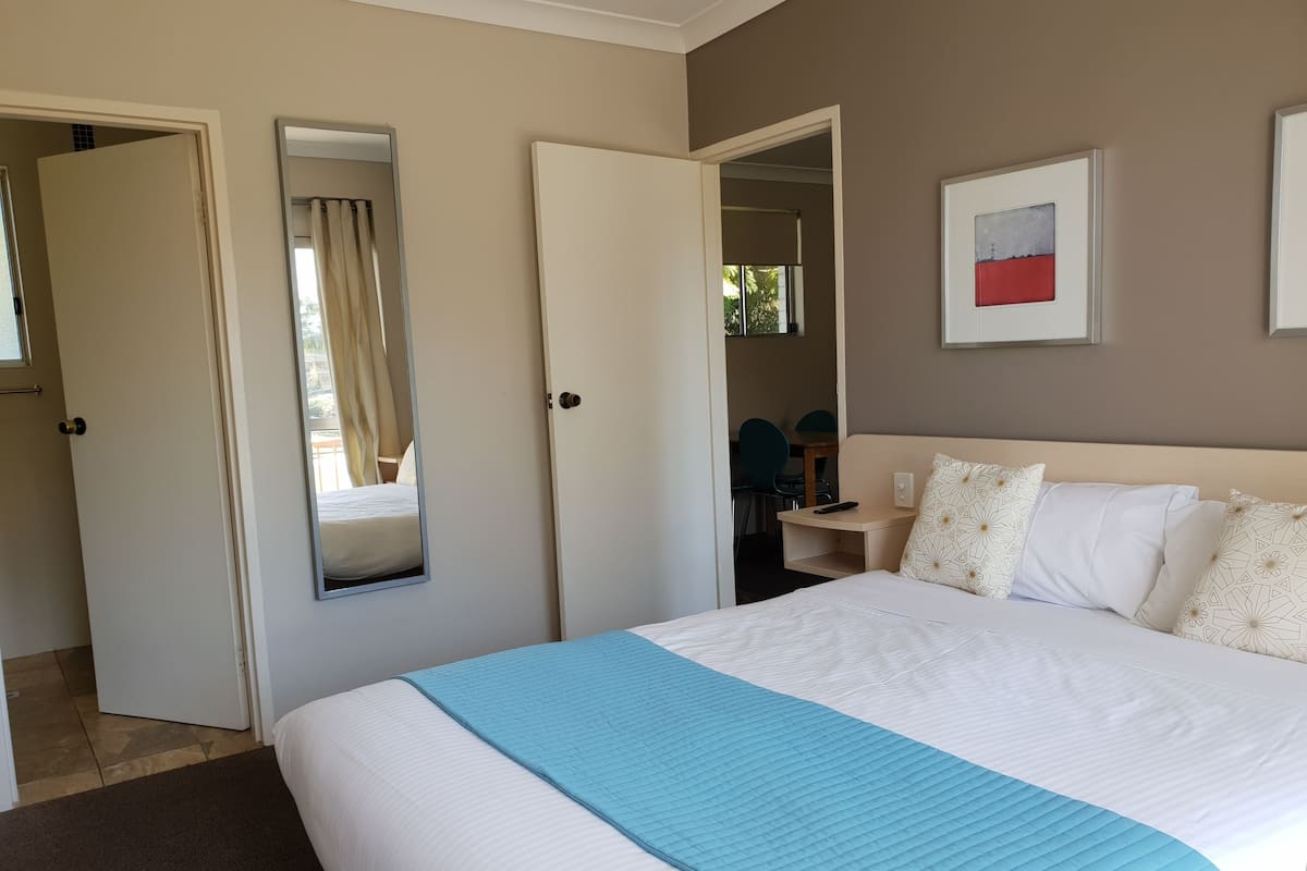 Quarto de hotel com uma cama grande, uma escrivaninha e uma cadeira. Há um espelho em uma parede e um quadro em outra.