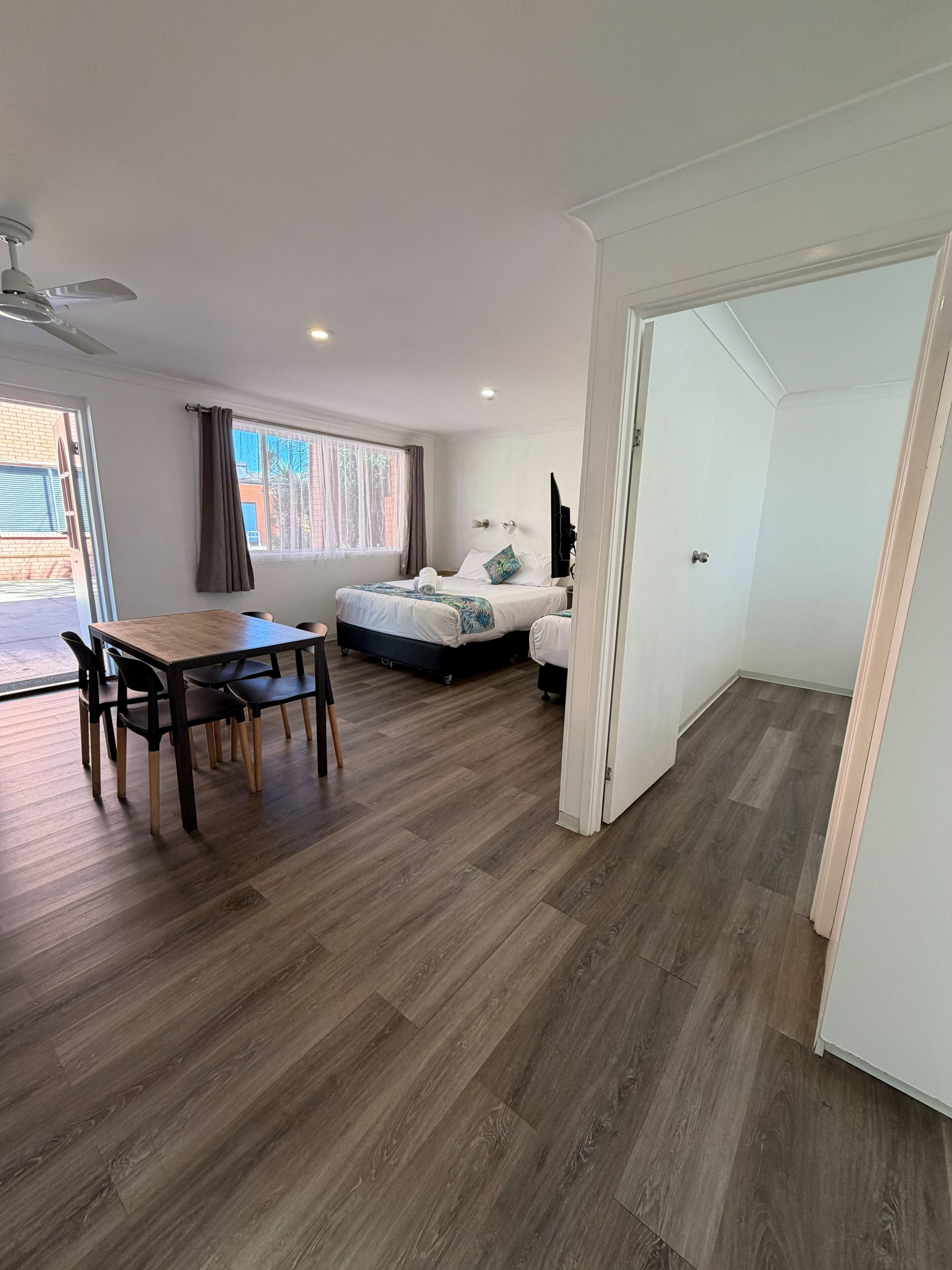 Quarto de hotel moderno com piso de madeira, cama com peseira branca, uma pequena mesa com cadeiras e uma janela com cortinas.