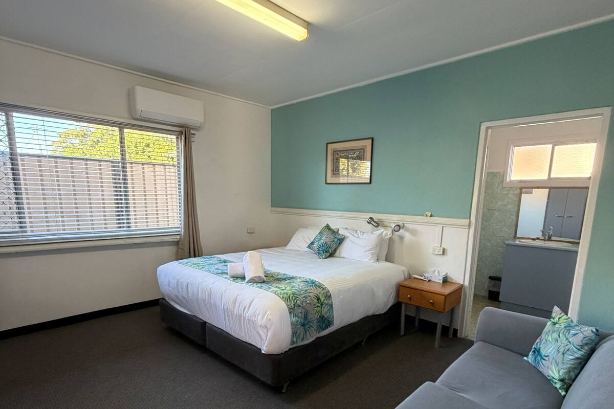 Quarto de hotel com cama, sofá, mesa de cabeceira, criadeira, espelho e janela com persianas.