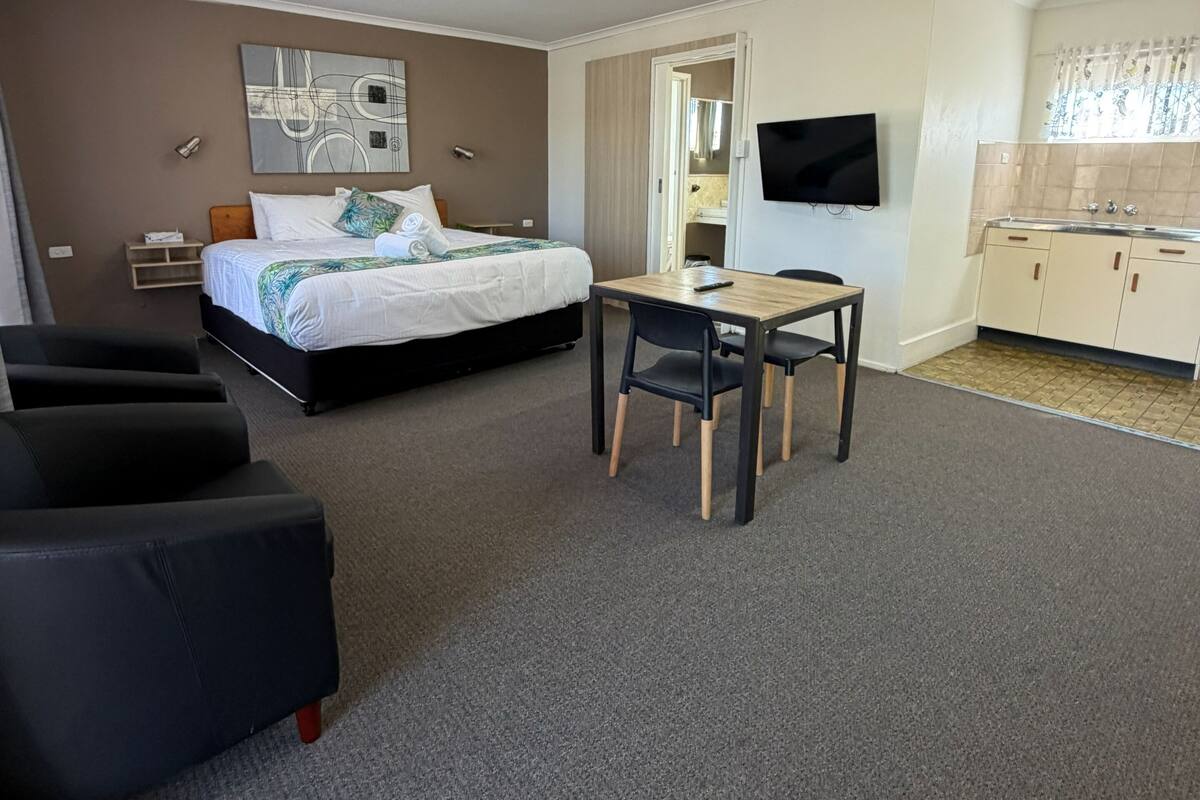 Quarto de hotel com uma cama, escrivaninha, cadeira, televisão e cozinha compacta.