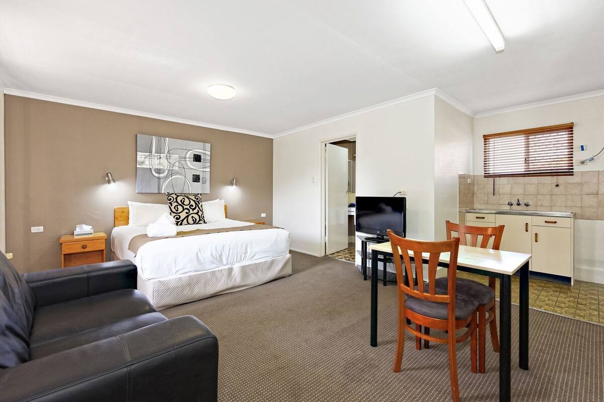 Quarto de hotel com cama, escrivaninha com televisão, mesa de jantar com cadeiras e cozinha compacta.