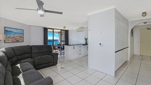 Uma sala de estar moderna com um sofá seccional cinza, um ventilador de teto e vista para o oceano através de janelas amplas.