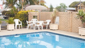 Piscina com cadeiras e mesa brancas, cercada por um muro de tijolos e plantas em vasos.
