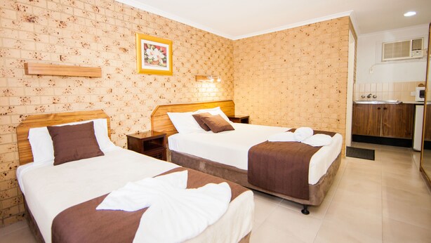 Quarto de hotel com duas camas, uma cozinha compacta e parede com padrão de tijolos.