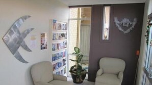 Um quarto com duas poltronas bege, uma estante com revistas, um objeto decorativo na parede e um vaso de planta.