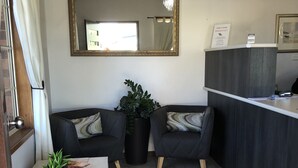 Uma área de recepção com um balcão escuro, um espelho grande, dois sofás pretos com almofadas estampadas, uma planta em vaso e uma pequena mesa com uma planta.