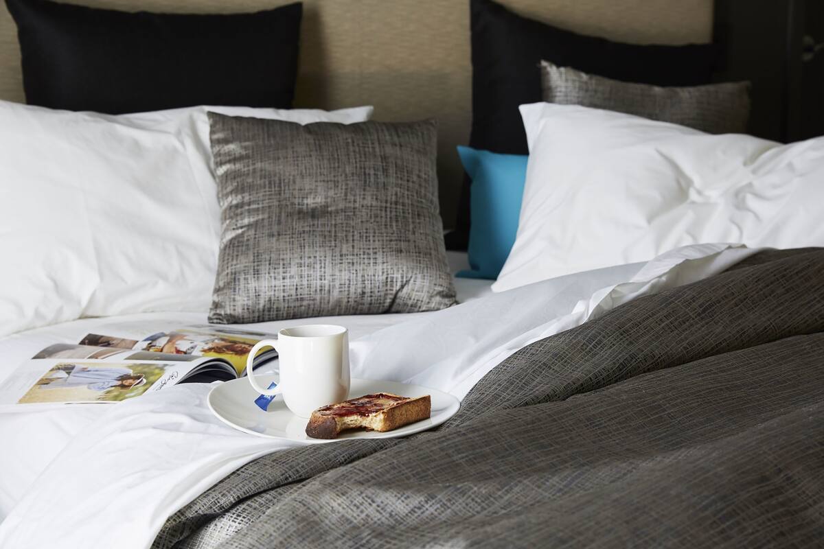 Cama bem arrumada com edredom cinza e branco, um travesseiro azul e um travesseiro branco. Uma fatia de torrada e uma caneca branca estão sobre a cama.