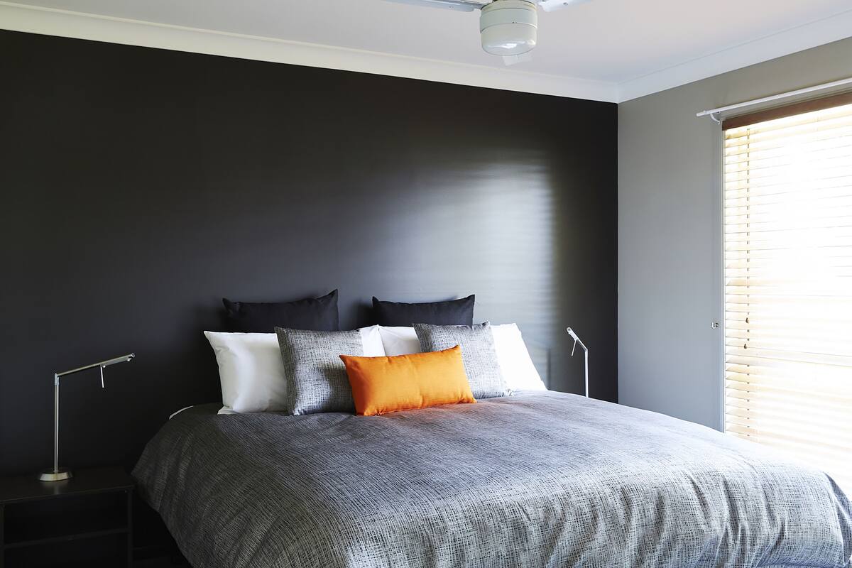 Quarto moderno com parede escura, cama com roupa de cama cinza e dois travesseiros, mesa de cabeceira com abajur e janela com persianas.