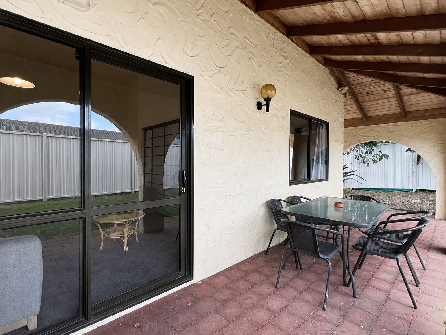 Um pátio externo com porta de vidro que dá para um quintal, uma mesa com cadeiras e um luminário fixado na parede.