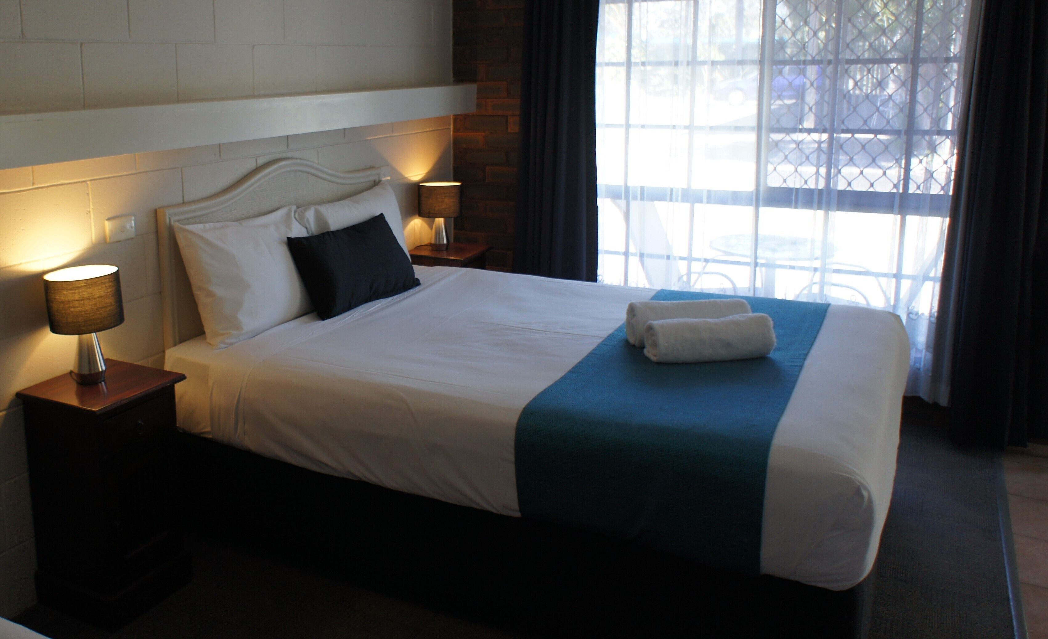Cama bem arrumada com roupa de cama branca e um detalhe azul, duas luminárias de cabeceira, uma janela com cortinas e vista para o exterior.