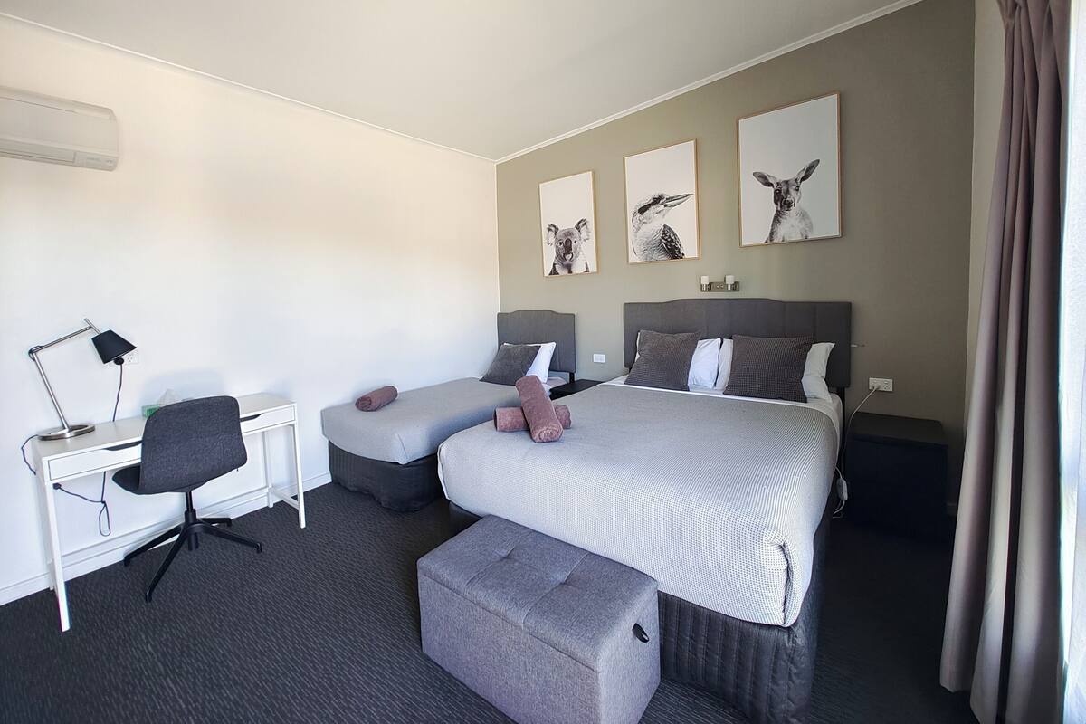 Quarto de hotel com escrivaninha, duas camas, uma cadeira e obras de arte nas paredes.
