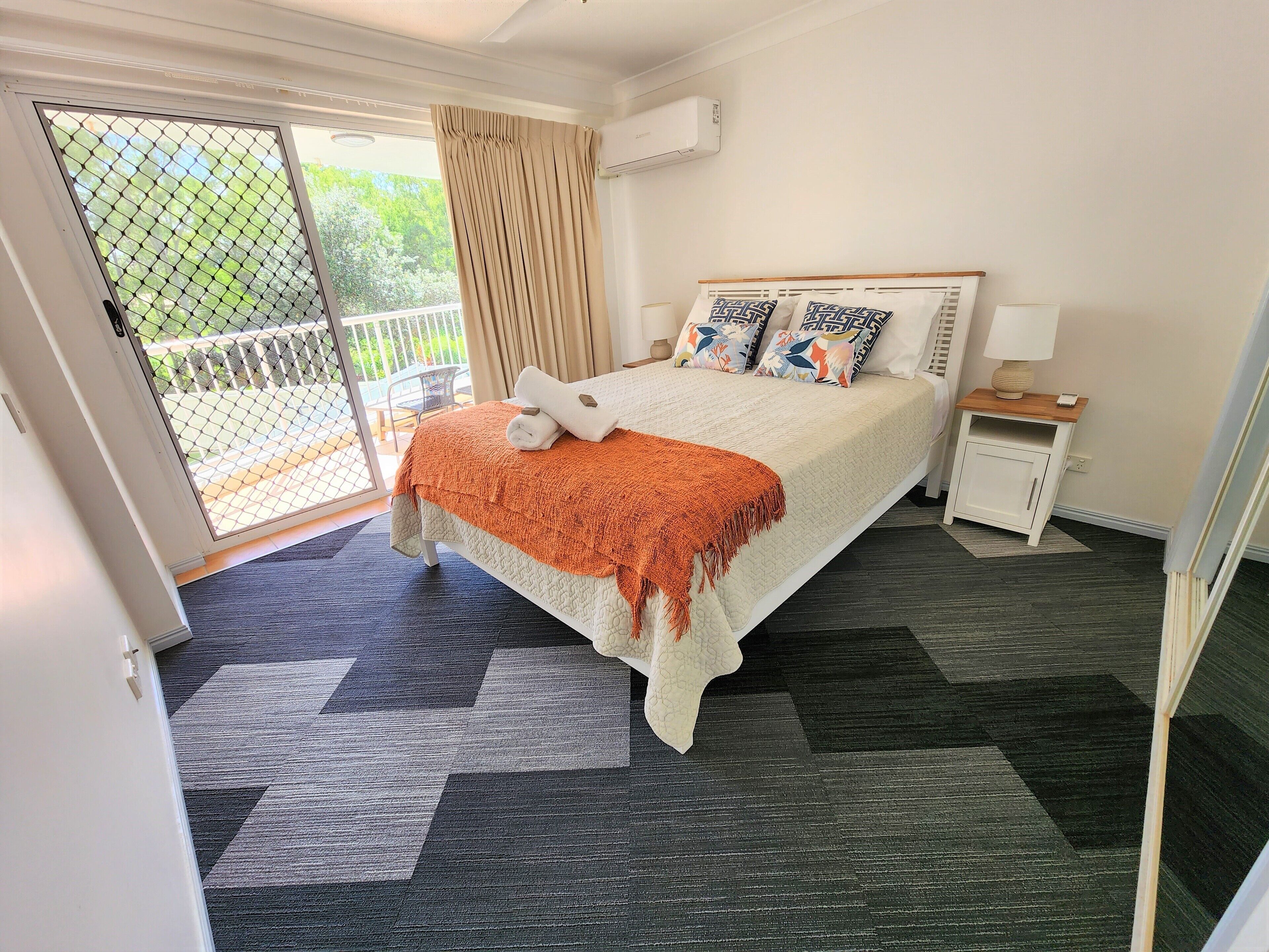 Quarto com cama, mesa de cabeceira, luminária, porta de vidro deslizante e vista para o verde lá fora.