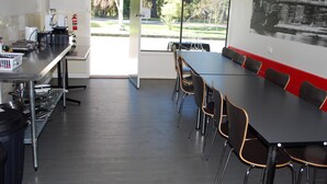 Uma sala de conferências com uma mesa comprida e cadeiras, uma área de cozinha com eletrodomésticos e uma janela grande com vista para árvores e para a paisagem urbana.