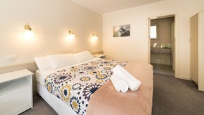Quarto de hotel com cama, mesa de cabeceira, luminária fixada na parede e banheiro visível por uma porta aberta.