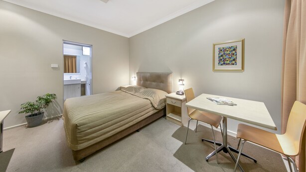 Quarto de hotel com cama, mesa de cabeceira, cadeira, escrivaninha e planta em vaso.