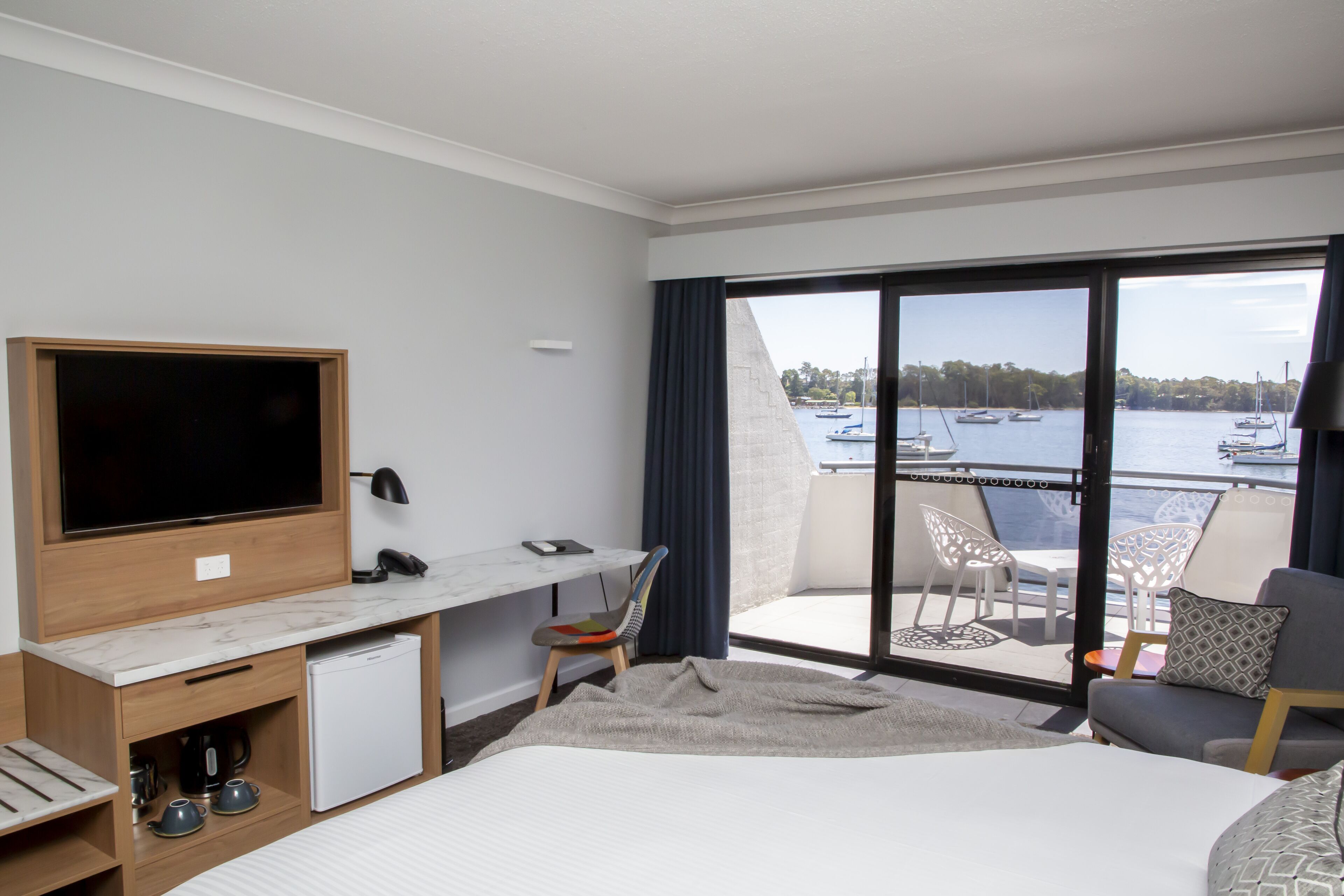Quarto de hotel com cama, escrivaninha, televisão, mini-frigorífico, cadeira e varanda com vista para barcos, além de mesa com cadeiras.