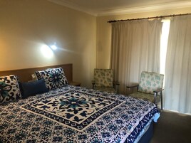 Quarto de hotel com uma cama grande, duas cadeiras, uma mesa de cabeceira e uma janela com cortinas.