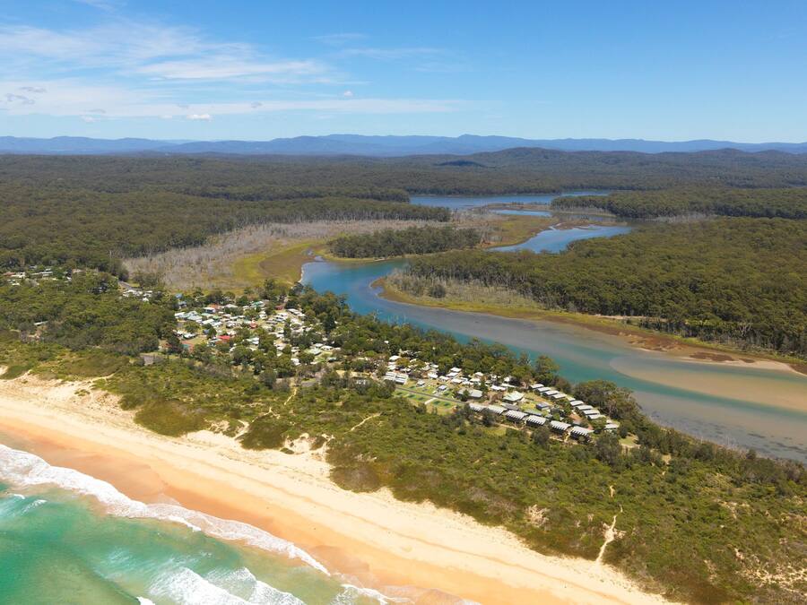 Vista aérea de uma área costeira com praia, rio e uma região arborizada com um conjunto de edifícios.