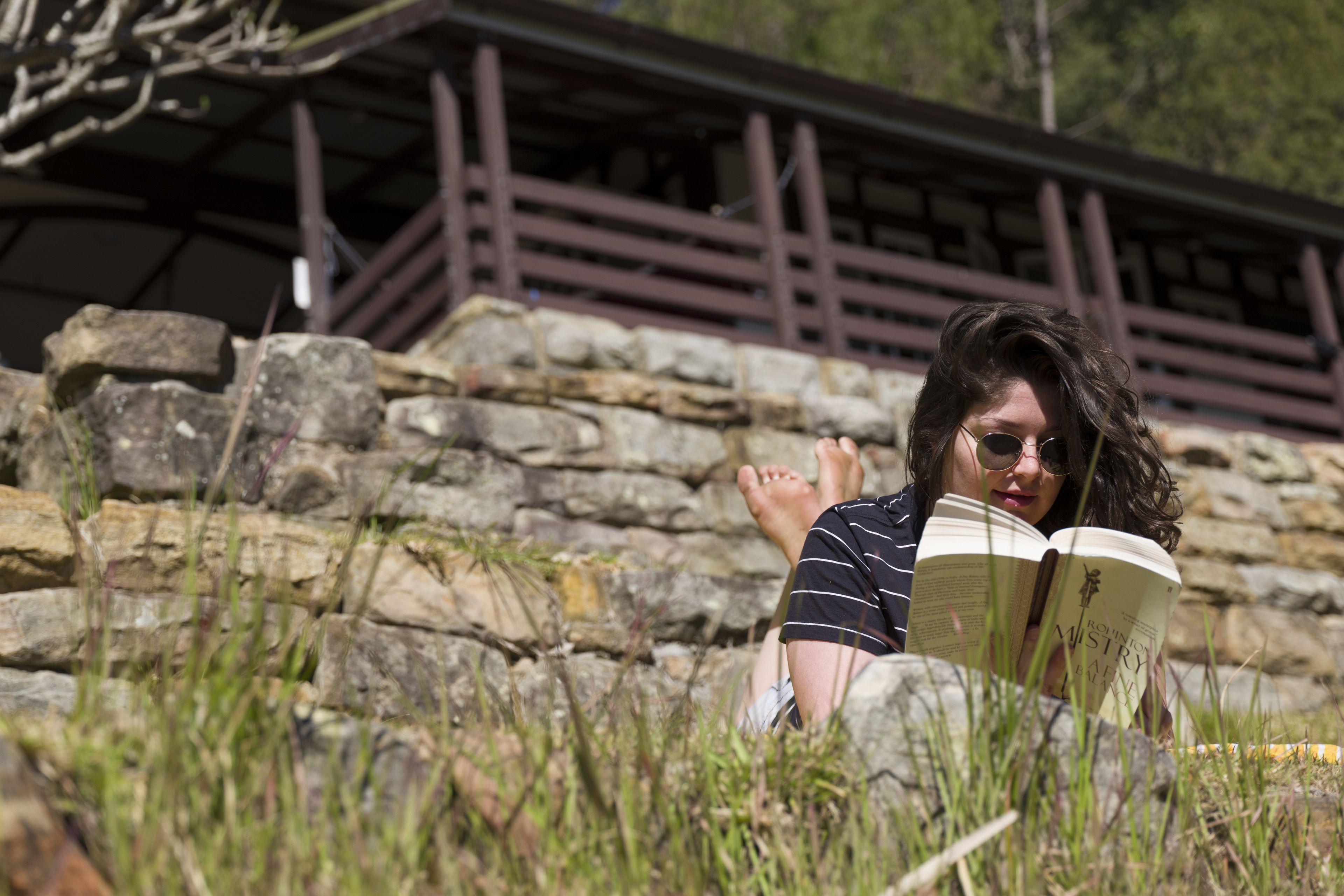 Uma pessoa lendo um livro ao ar livre, com um muro de pedra e estrutura de madeira ao fundo.