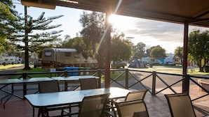Uma área de estar ao ar livre com mesas e cadeiras, uma estrutura coberta e vista para uma área de camping com veículos recreativos e árvores.