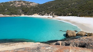 Paisagem costeira com mar azul-turquesa límpido, praia de areia branca e afloramentos rochosos.