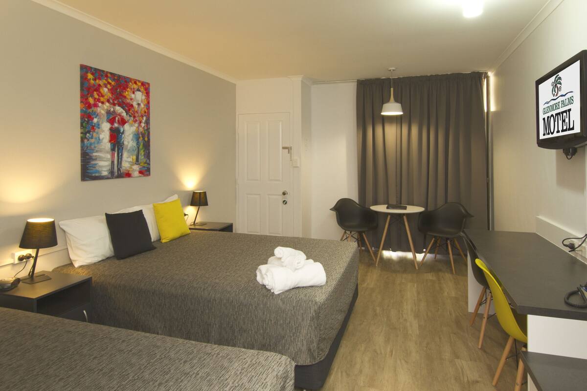 Quarto de hotel com cama, duas mesas de cabeceira, escrivaninha, cadeira, um quadro na parede e uma placa indicando que é um motel.
