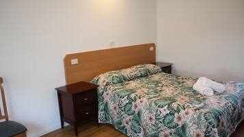 Quarto com cabeceira de madeira, uma cama de solteiro com colcha floral, mesa de cabeceira, cadeira de madeira e uma pequena mesa.