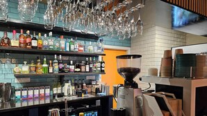 Uma área de bar com prateleiras de garrafas de bebidas, uma máquina de café e copos pendurados.