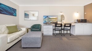 Quarto de hotel com sofá branco, poltrona verde, banqueta cinza, escrivaninha com duas cadeiras, um quadro grande, um espelho e uma cozinha compacta.