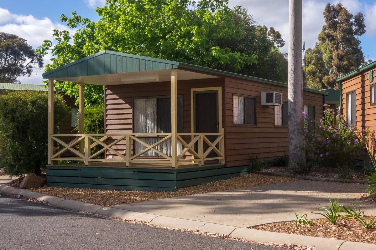 Uma cabana pequena e térrea, com varanda, rodeada por árvores e um céu claro.