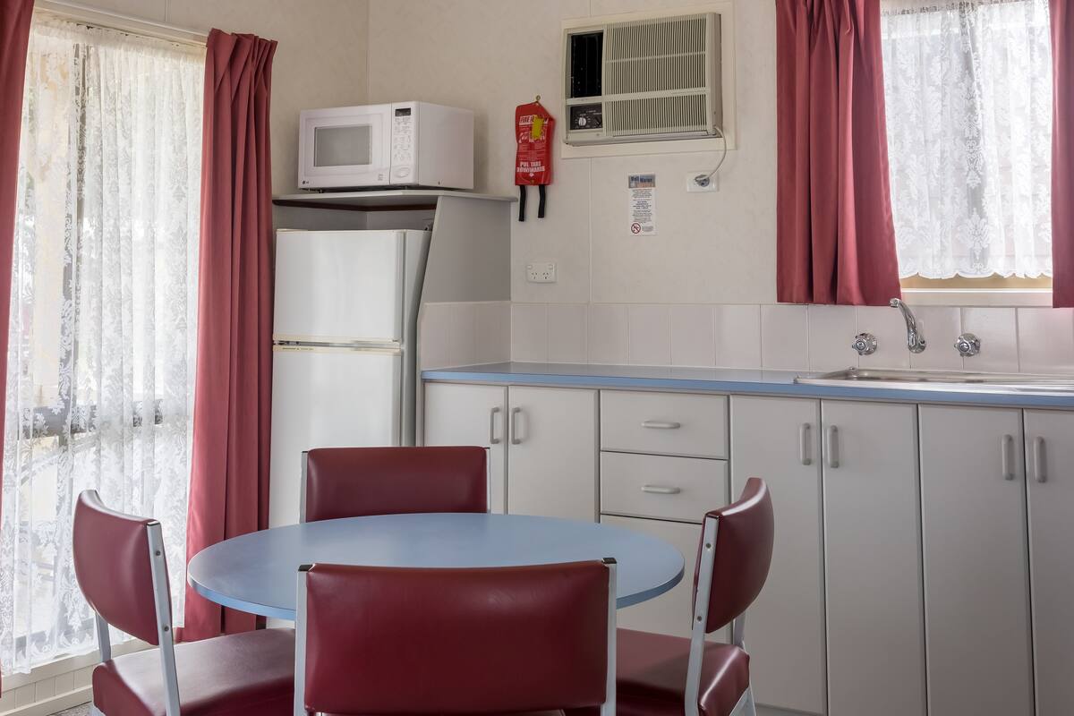 Cozinha compacta com micro-ondas, geladeira e pia. Há uma mesa com quatro cadeiras e uma janela com cortinas.