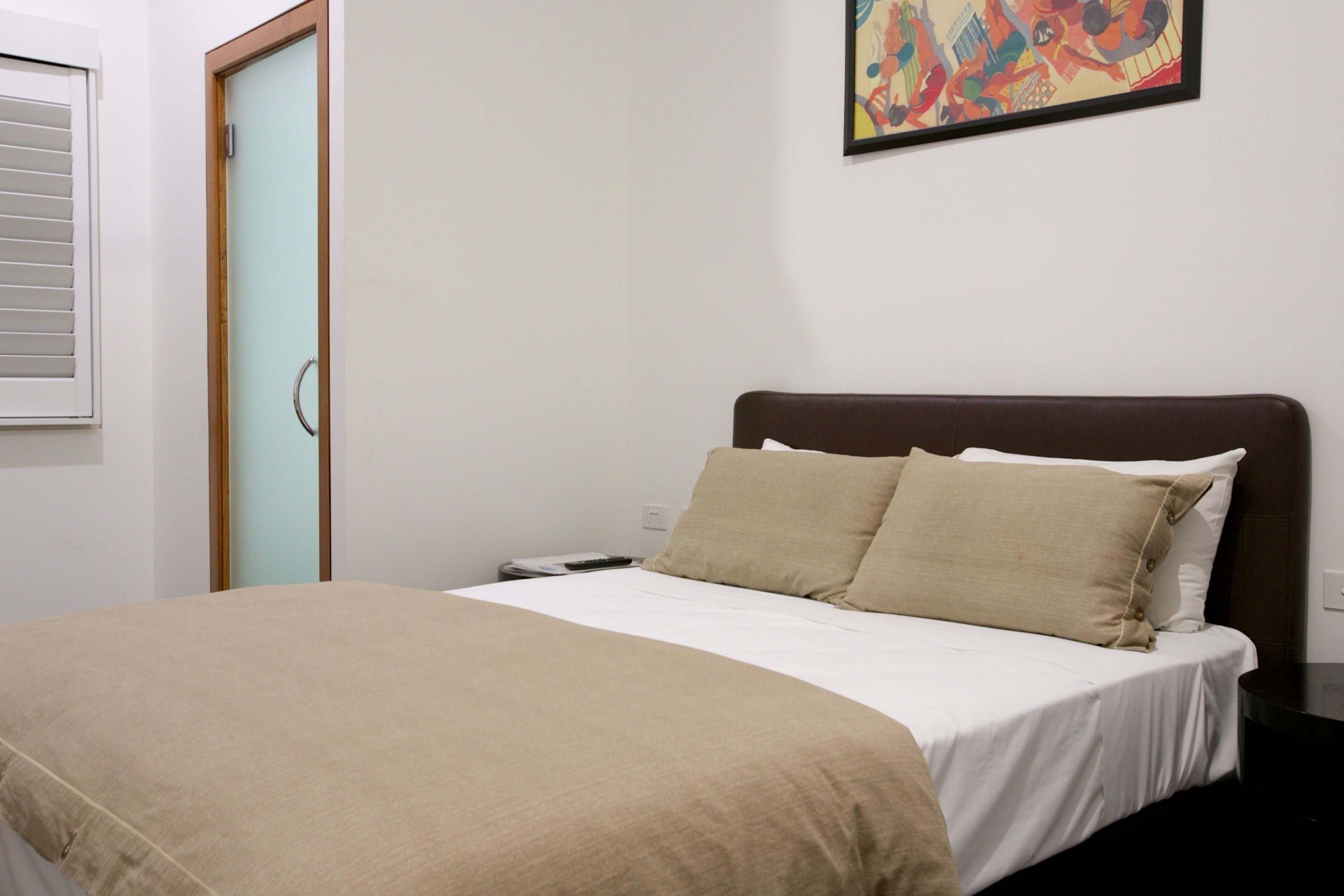 Cama bem arrumada com cabeceira marrom, manta bege e dois travesseiros.