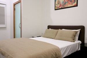 Cama bem arrumada com cabeceira marrom, manta bege e dois travesseiros.