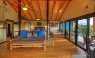 Quarto espaçoso com uma cama grande, piso de madeira, ventilador de teto e vista para o exterior por uma porta de vidro deslizante.