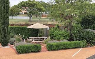 Uma área de pátio com mesa e guarda-sol, rodeada por sebes bem cuidadas e diversas plantas.