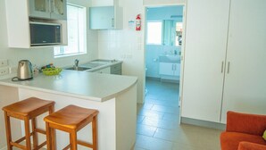 Cozinha moderna com armários brancos, micro-ondas e chaleira. Há banquetas de madeira e uma cadeira vermelha. A cozinha dá para um banheiro com pia e vaso sanitário.