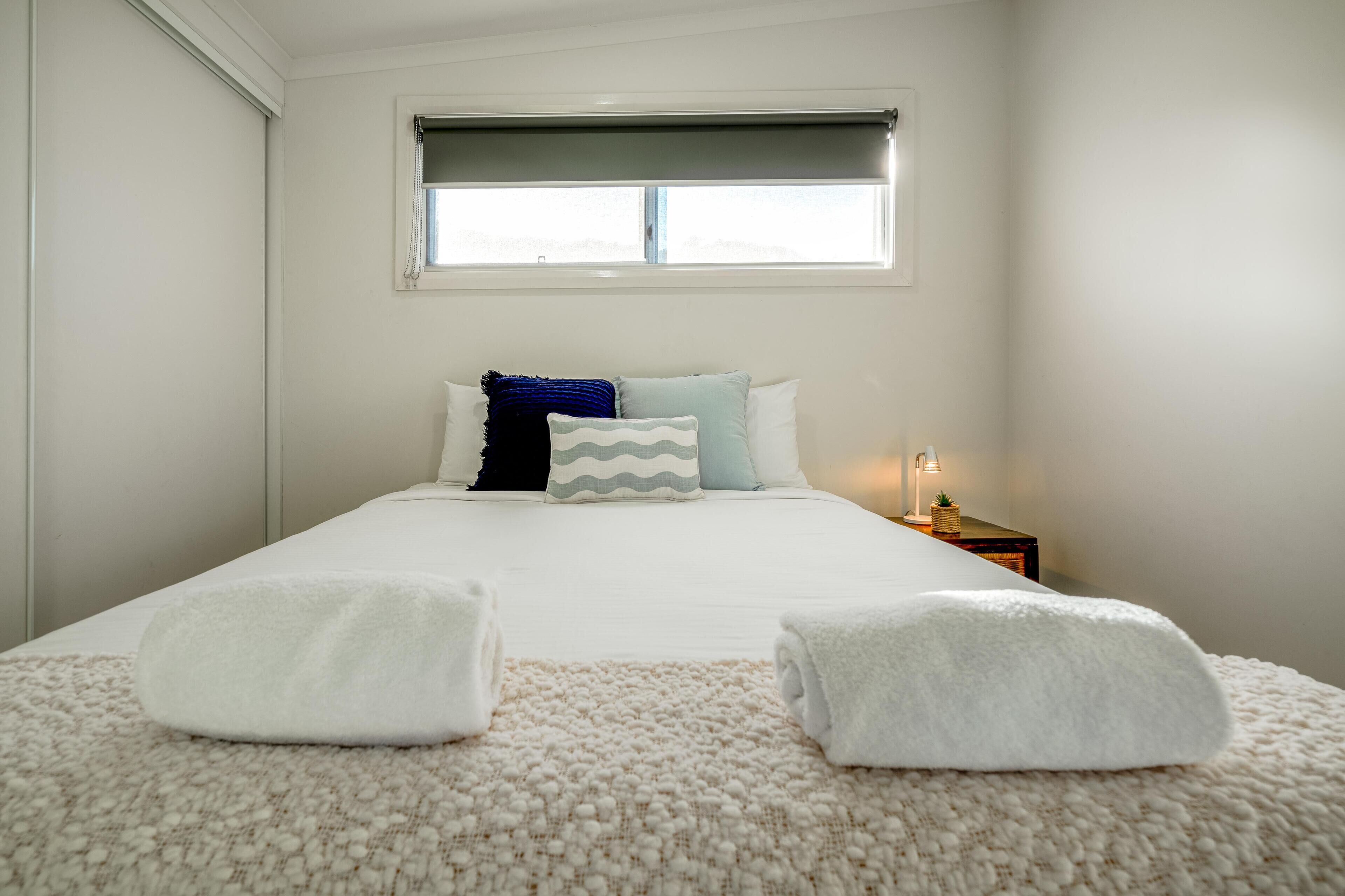 Cama bem arrumada com lençóis brancos e duas toalhas, mesa de cabeceira com abajur, e janela com persiana.
