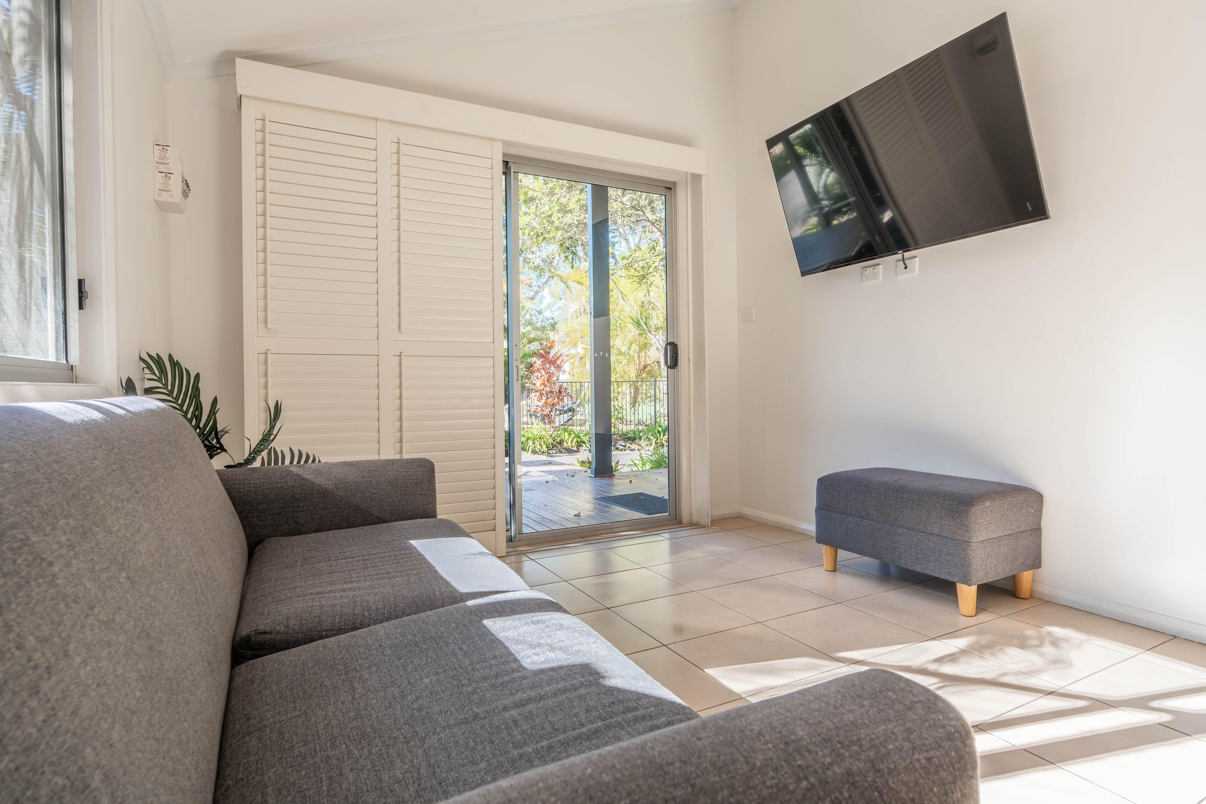 Uma sala de estar moderna com um sofá cinzento, um banquinho cinzento, uma TV de tela plana na parede e uma porta de vidro deslizante para uma área externa.