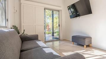 Uma sala de estar moderna com um sofá cinzento, um banquinho cinzento, uma TV de tela plana na parede e uma porta de vidro deslizante para uma área externa.