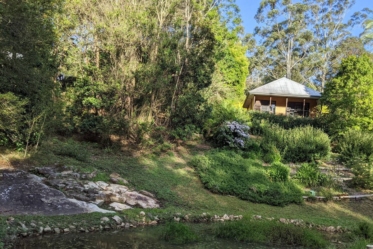 Uma pousada rodeada por floresta densa, um pequeno riacho e um jardim bem cuidado.
