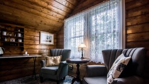 Um quarto acolhedor com paredes de madeira, duas poltronas estofadas, uma mesa pequena com abajur, um quadro na parede e uma estante com objetos para chá.