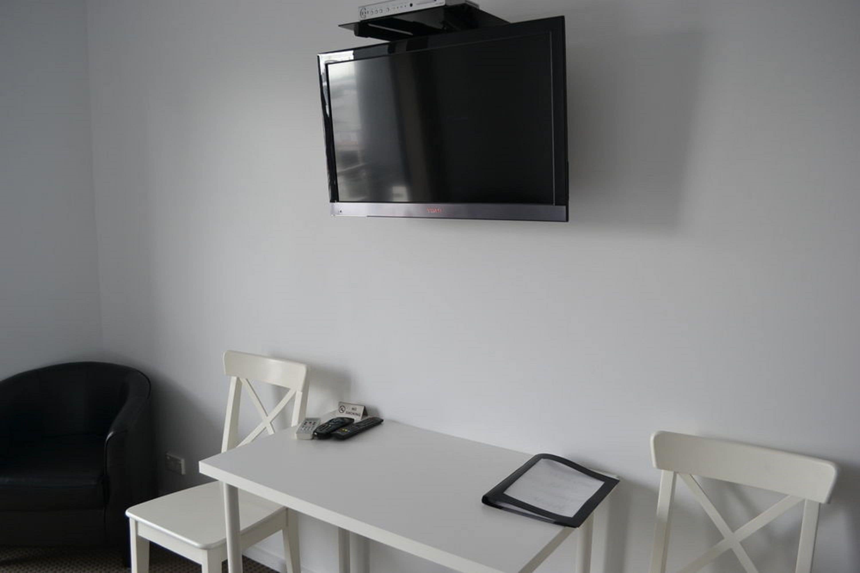 Quarto com televisão de tela plana na parede, escrivaninha branca com cadeira, controle remoto e tablet.